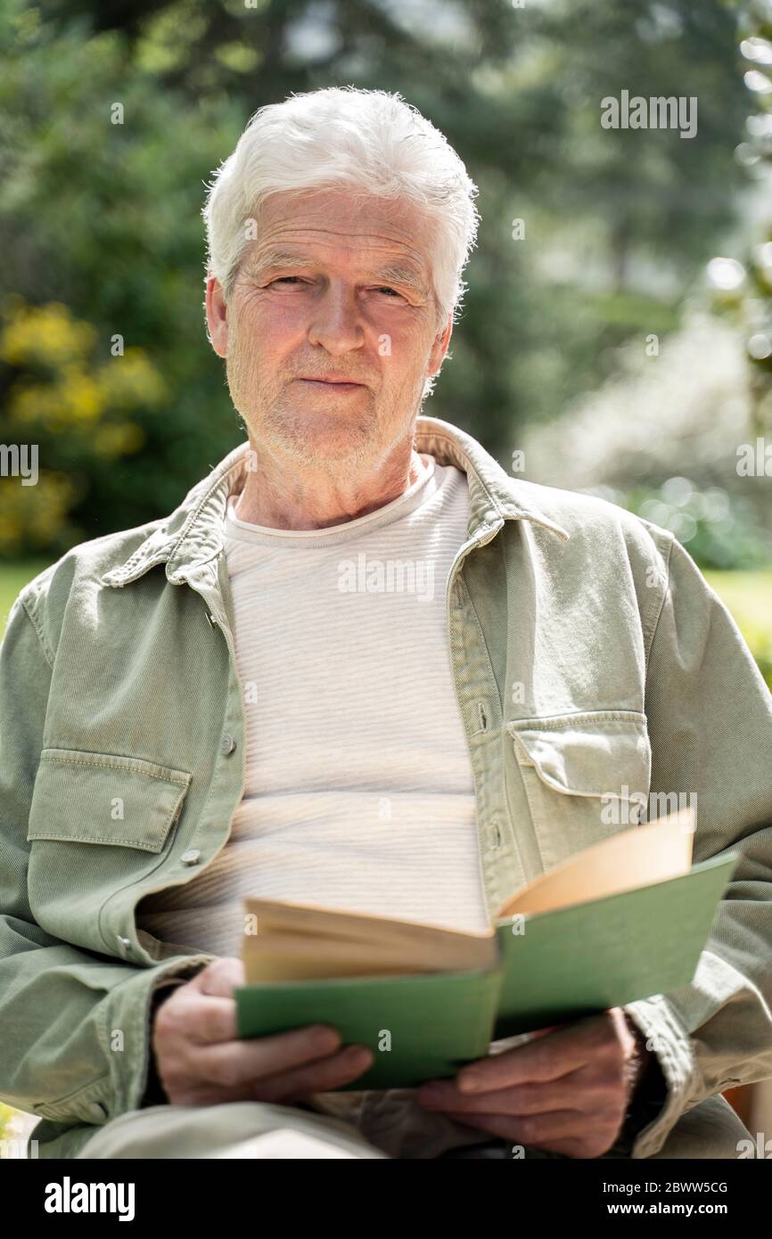 Portrait of senior man reading book while sitting in back yard on sunny ...