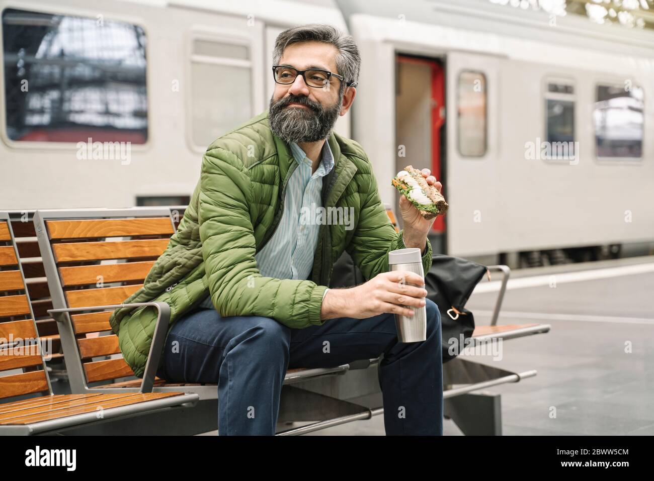 Man sitting on a bench at the train station with sandwich and hot drink ...
