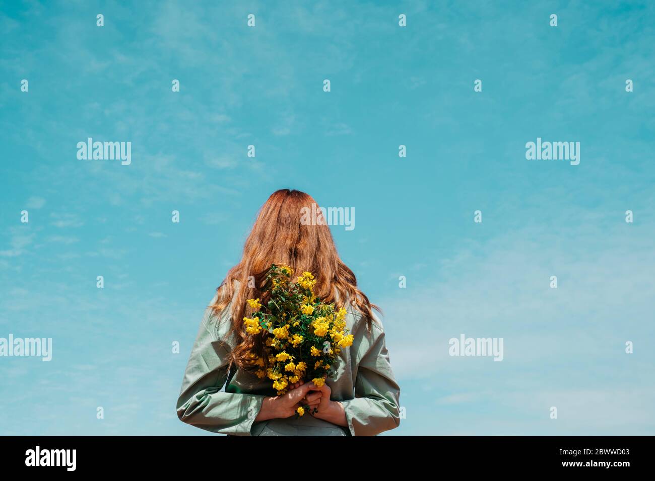 Back view of redheaded young woman against sky holding bunch of flowers ...