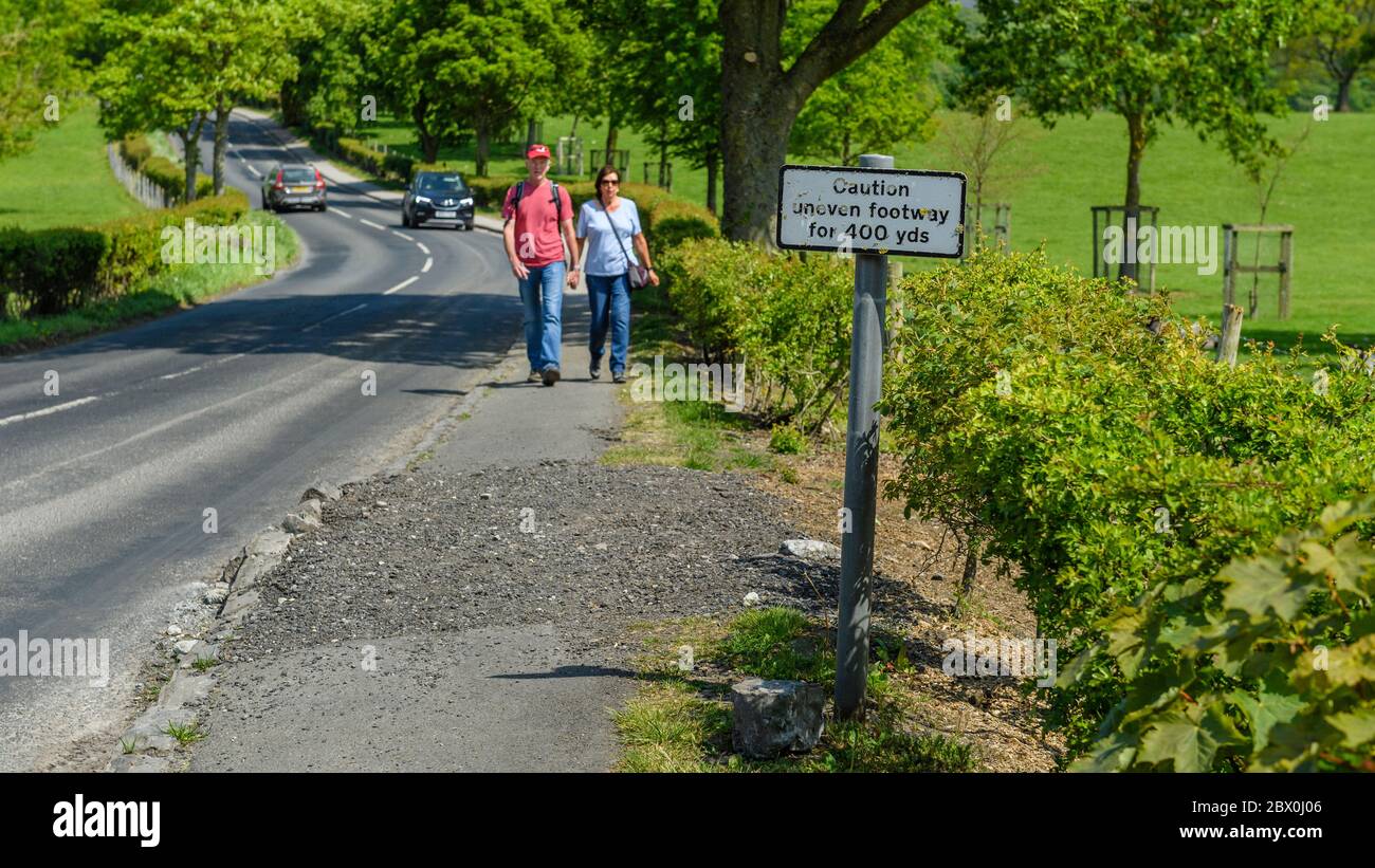 2 people walking by country lane, approaching 'Caution uneven footway ...