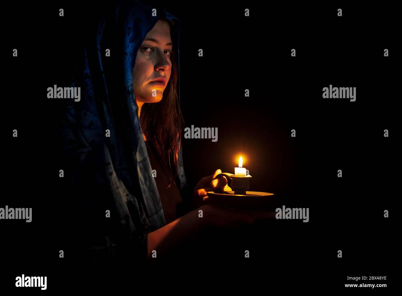 Sad young woman with candle and blue headscarf against black background ...