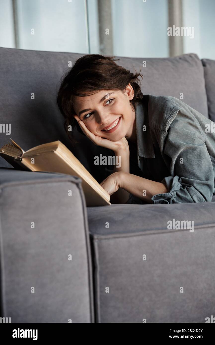 Image of brunette cute pleased woman reading book and smiling while ...
