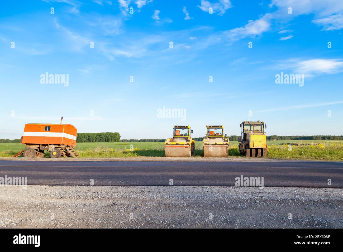 Three yellow pavers next to a green field and trees and a blue sky with ...