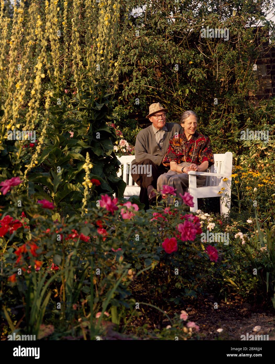Elderly couple surrounded by flowers sitting on white bench in summer ...
