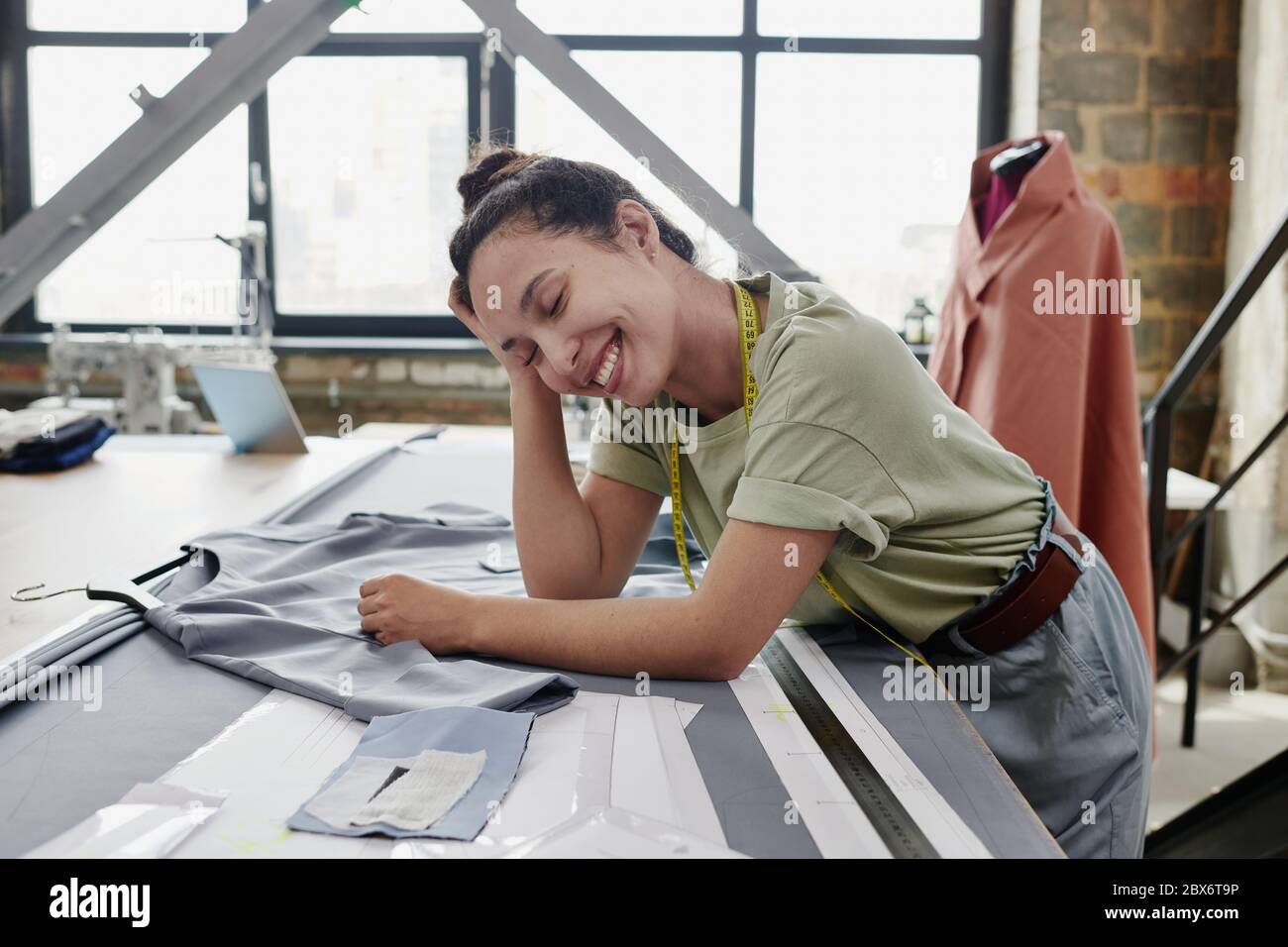 Young cheerful tailor or designer of clothes in casualwear leaning on ...