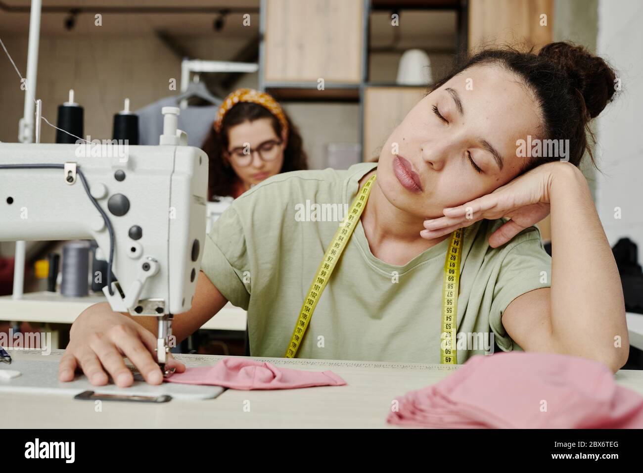 Young tired seamstress with measuring tape on her neck sitting by ...