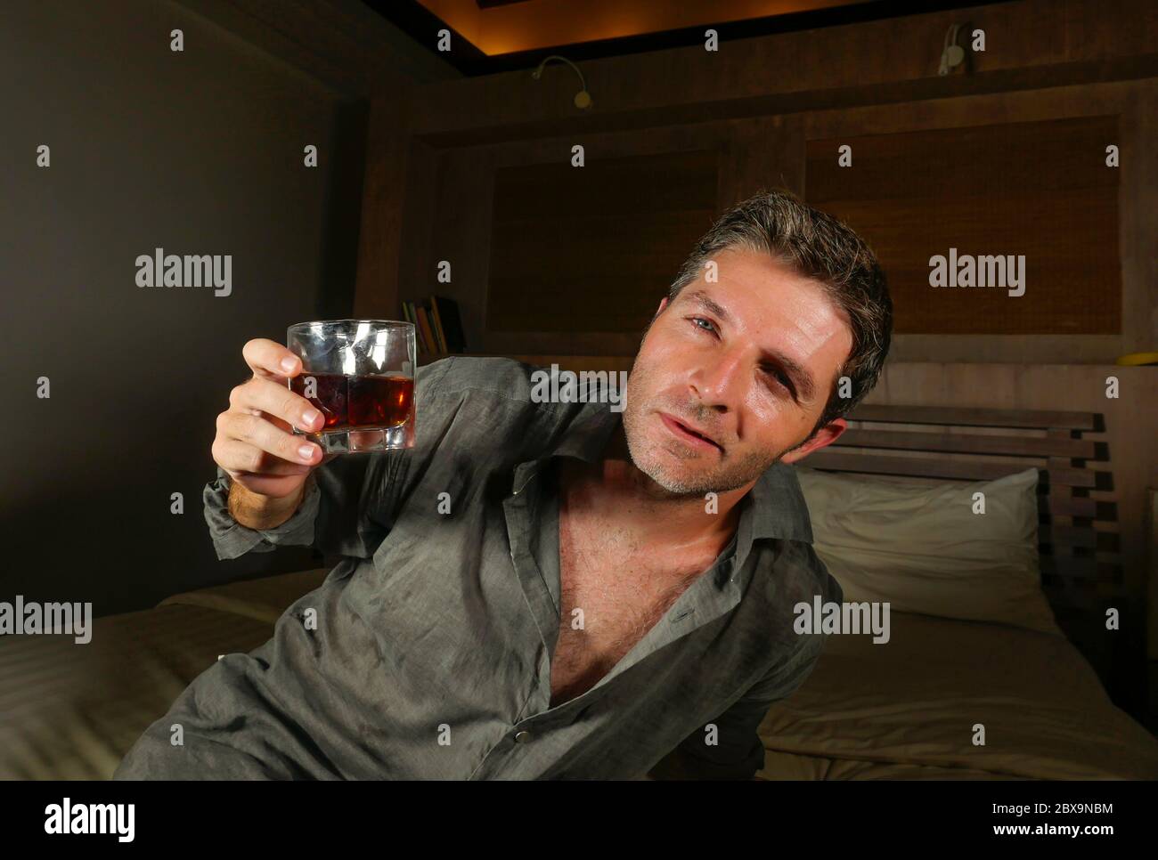 young messy and wasted drunk man drinking whiskey glass at home sitting on bed hammered and ...