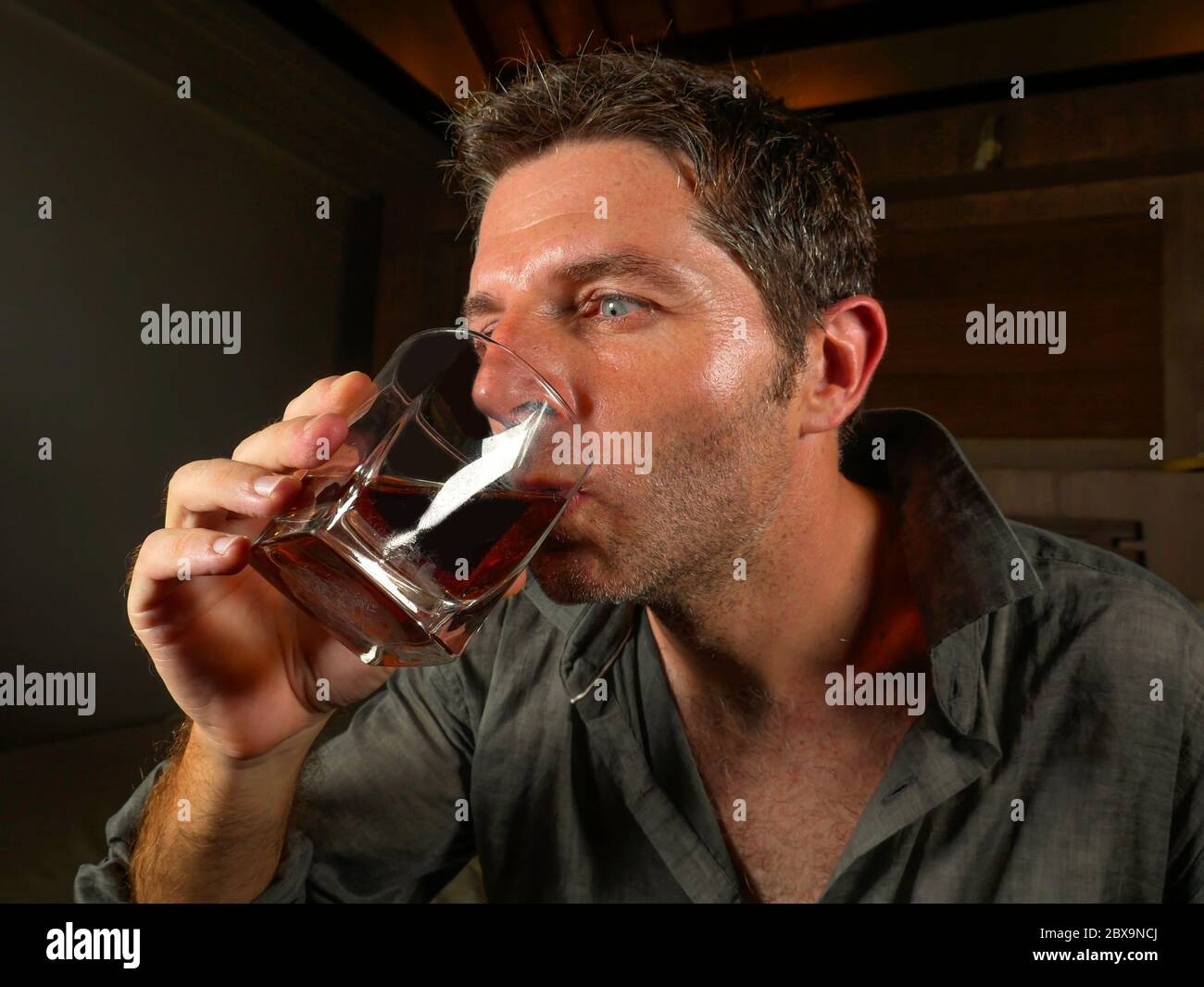 young messy and wasted drunk man drinking whiskey glass at home sitting on bed hammered and ...