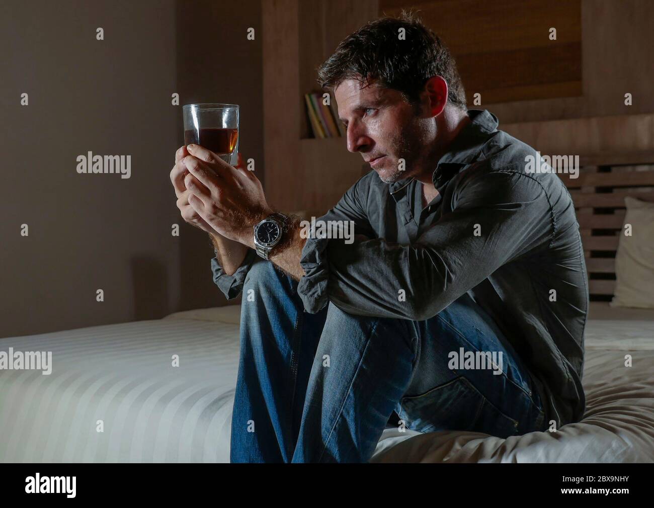 young messy and wasted drunk man drinking whiskey glass at home sitting on bed hammered and ...