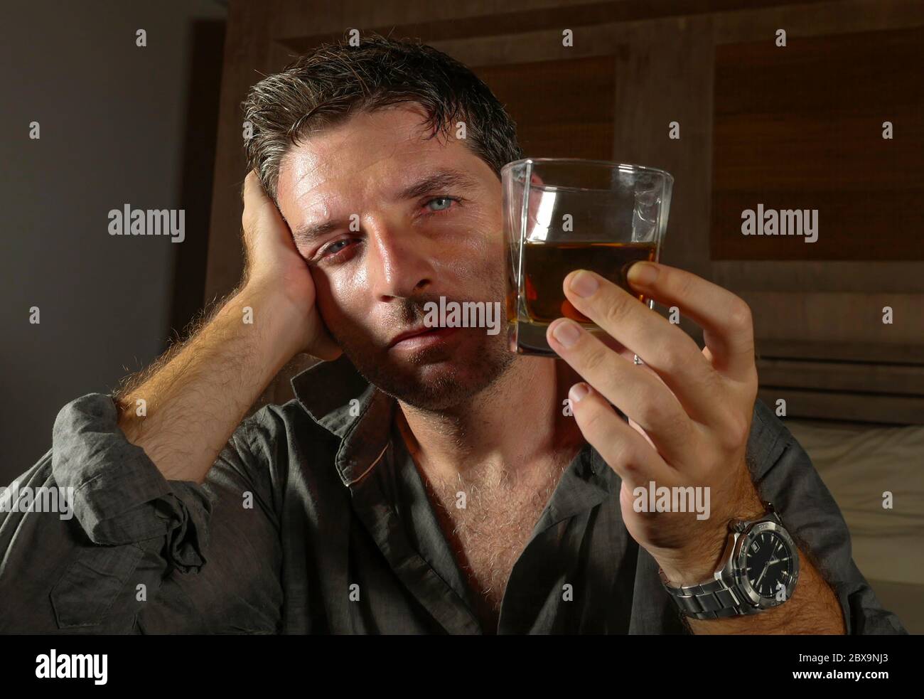 young messy and wasted drunk man drinking whiskey glass at home sitting on bed hammered and ...