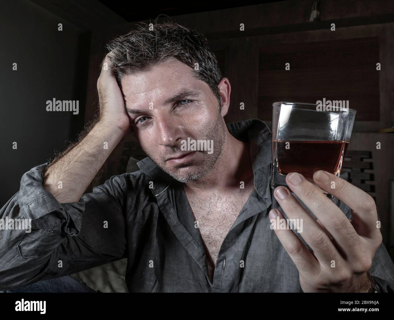 young messy and wasted drunk man drinking whiskey glass at home sitting on bed hammered and ...