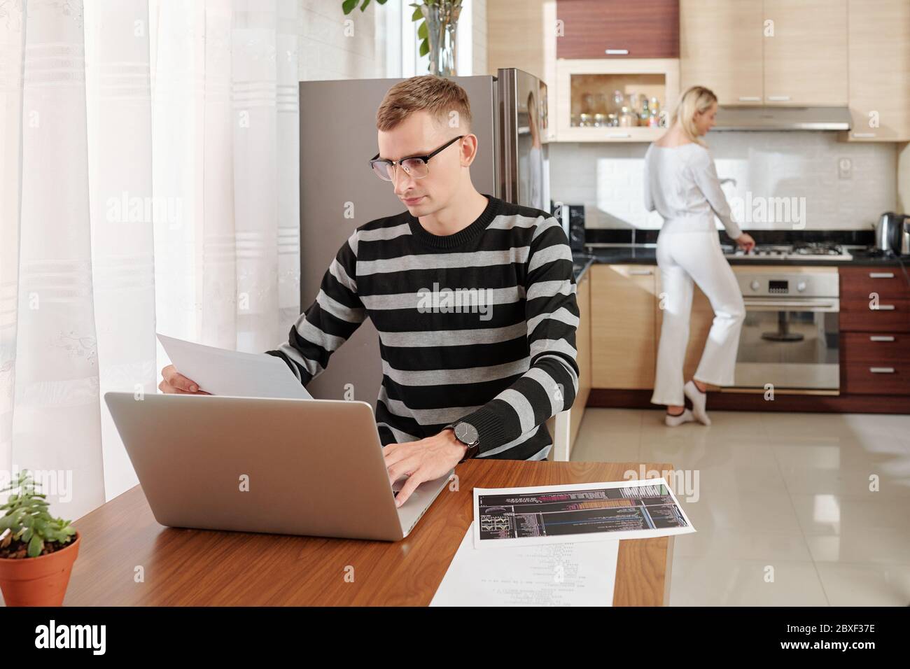 Software developer coding at kitchen table when his wife cooking dinner ...