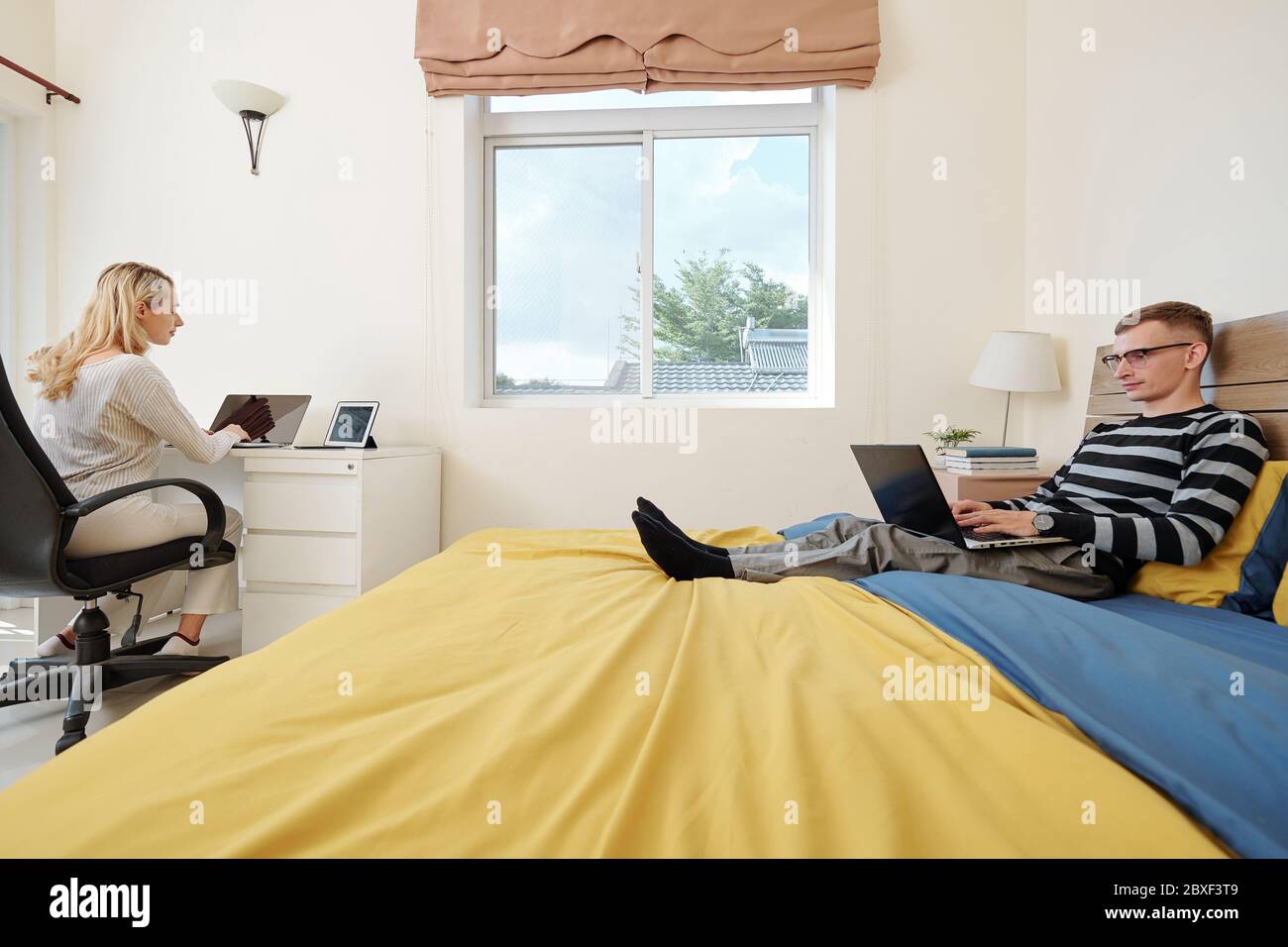 Young man sitting on bed and programming on laptop when his wife ...