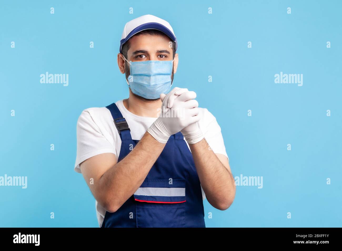 Optimistic worker in overalls, mask and gloves keeping hands together ...