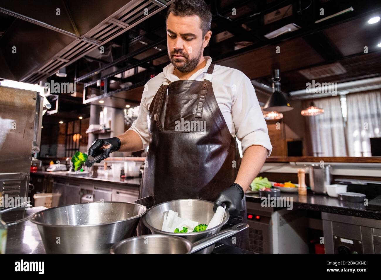 Middle-aged bearded chef in apron and gloves standing at stove and ...