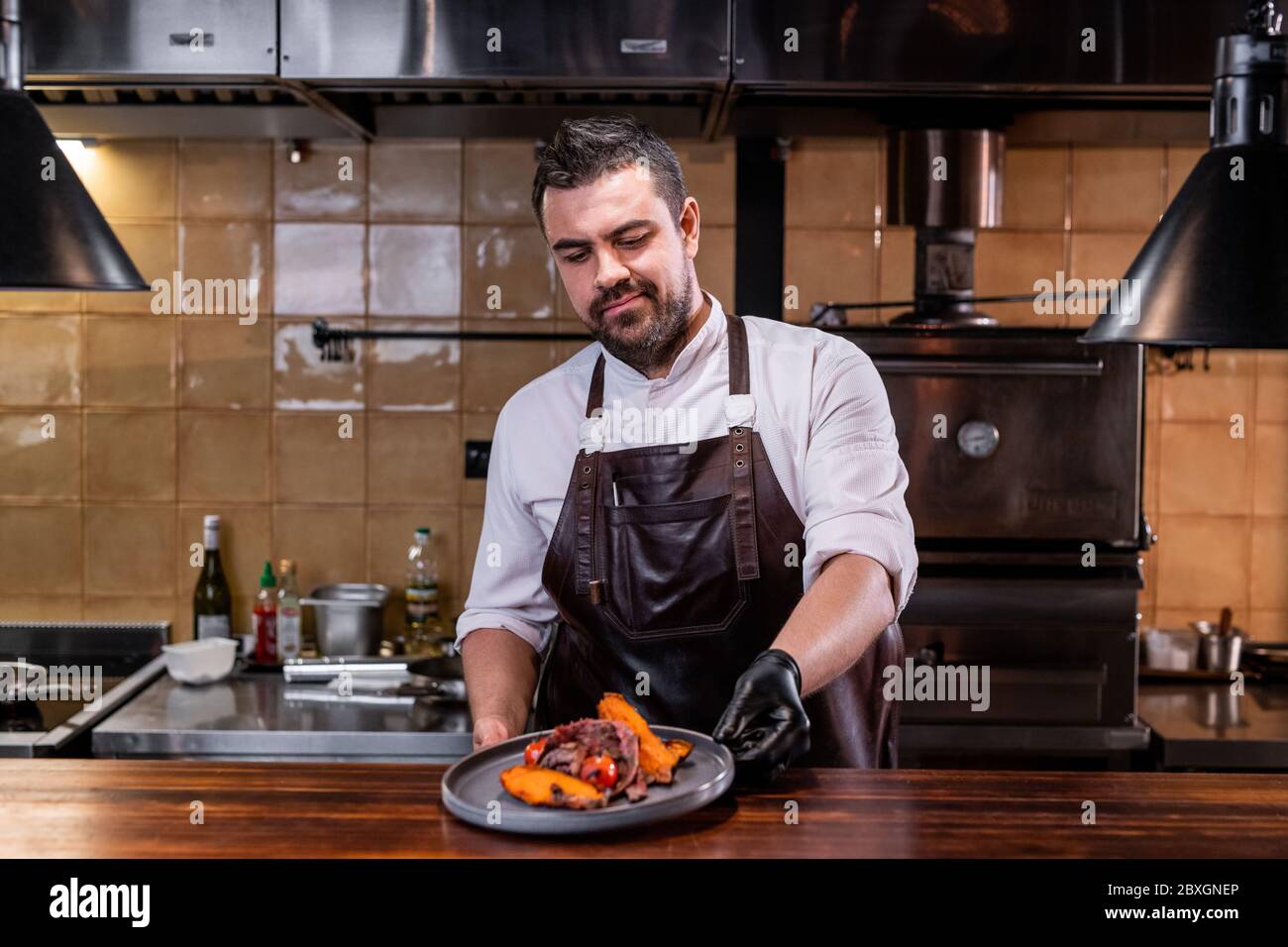 Bearded chef in leather apron putting prepared dish on counter in ...