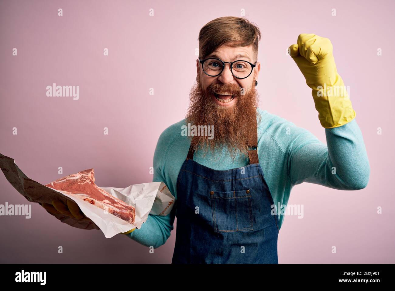 Redhead Irish butcher man with beard holding raw beef steak over pink ...