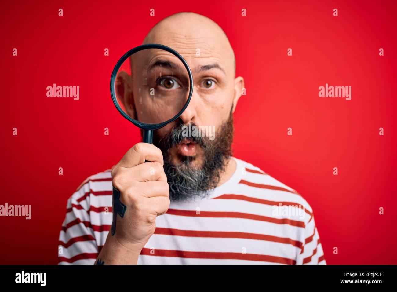 Handsome detective bald man with beard using magnifying glass over red ...