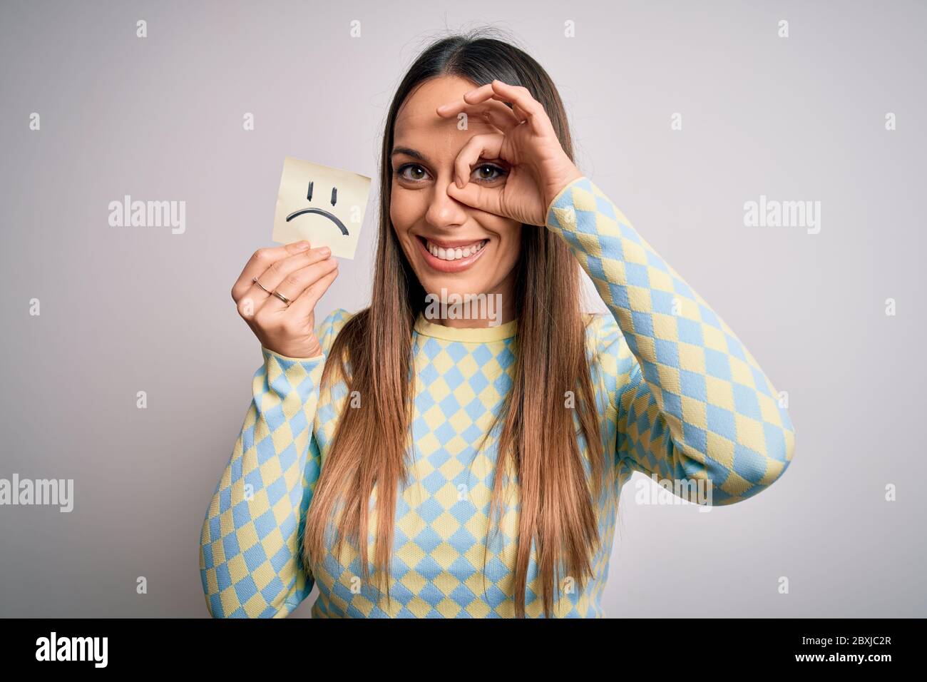 Young blonde woman holding sticky paper note with sad face emoticon ...