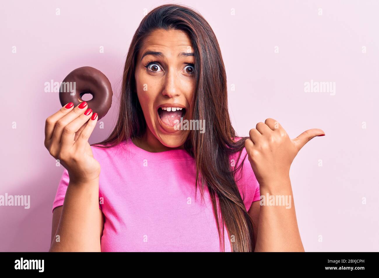 Young beautiful brunette woman holding chocolate donut pointing thumb ...