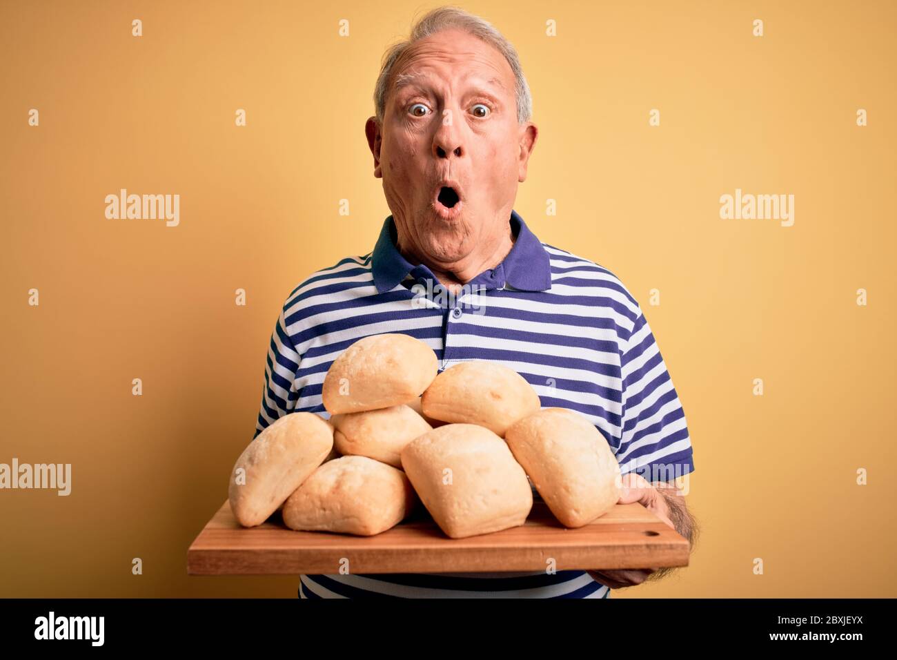 Senior grey haired baker man holding fresh homemade bread over yellow ...