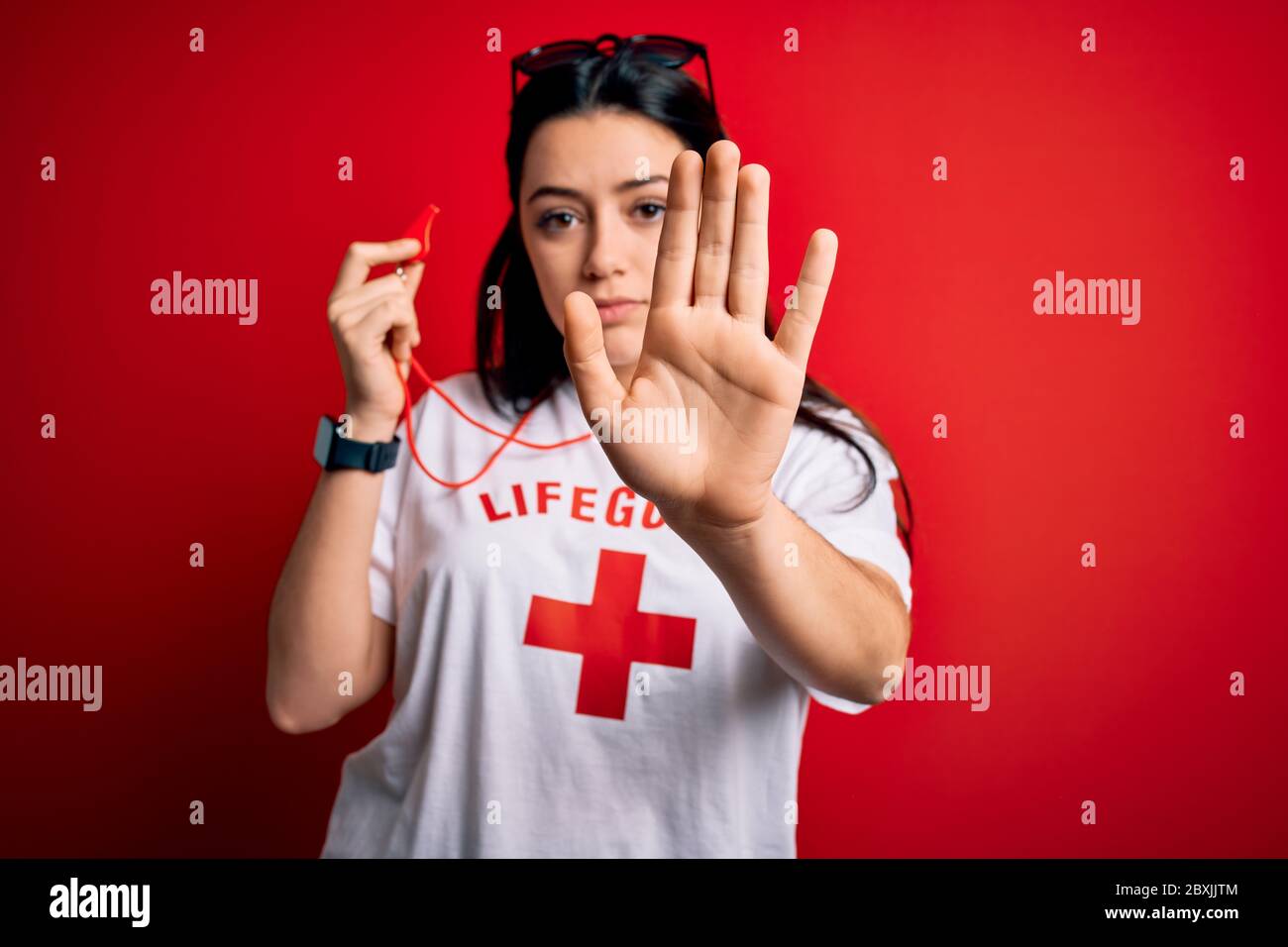 Young lifeguard woman wearing guard equipement holding whistle over red ...