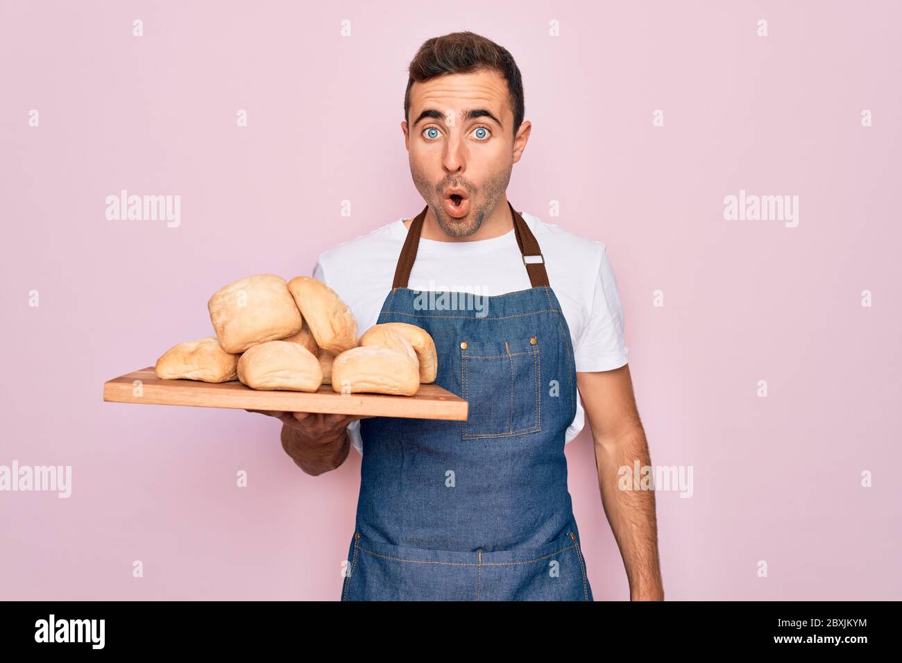 Young handsome baker man with blue eyes wearing apron holding tray with ...