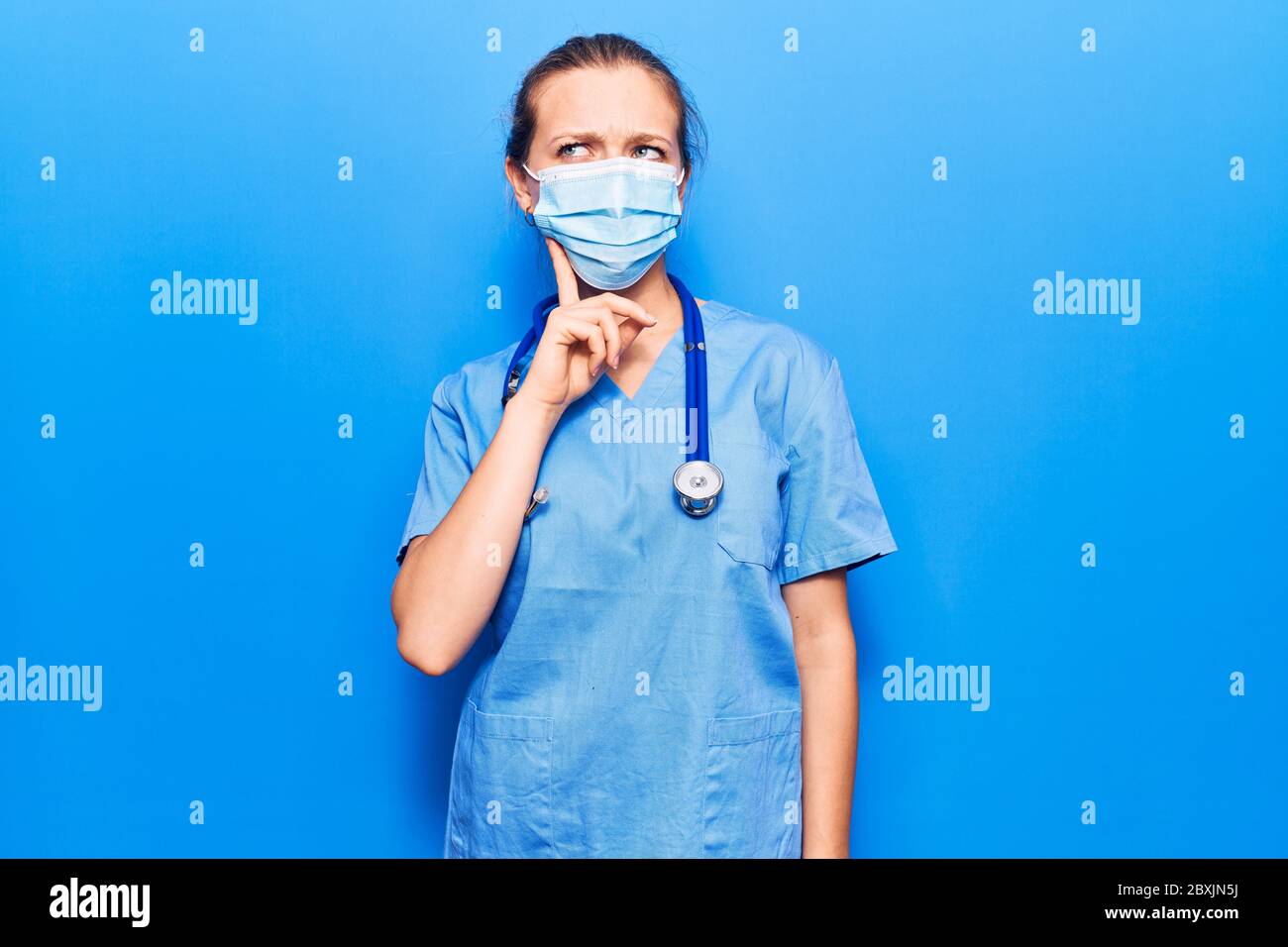 Young blonde woman wearing doctor uniform and medical mask thinking ...
