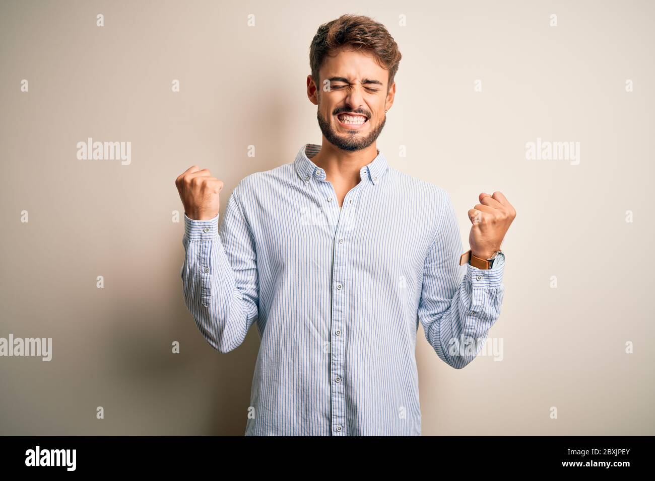 Young handsome man with beard wearing striped shirt standing over white ...