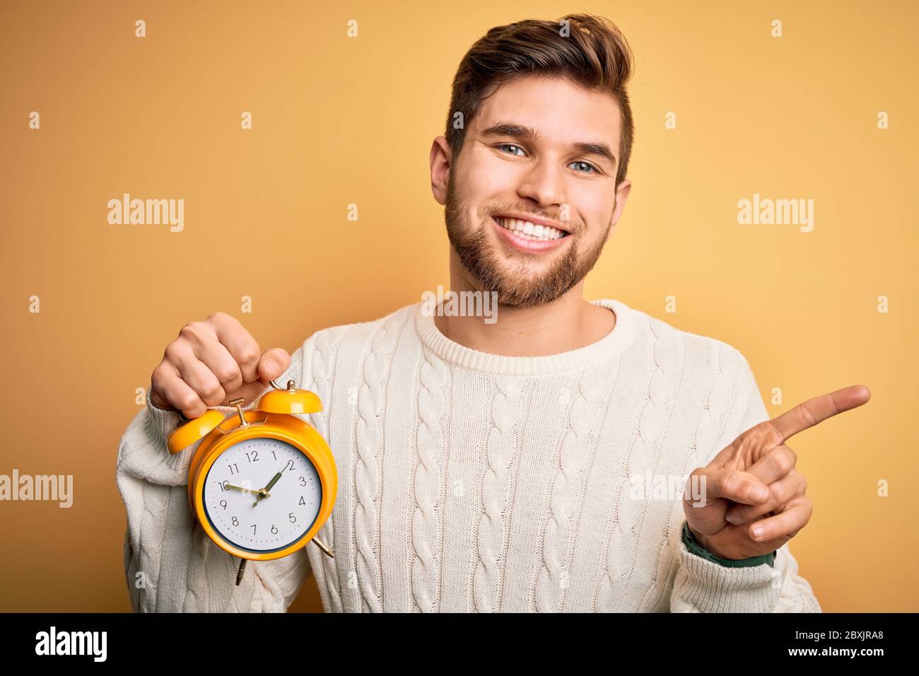 Young blond man with beard and blue eyes holding alarm clock over ...