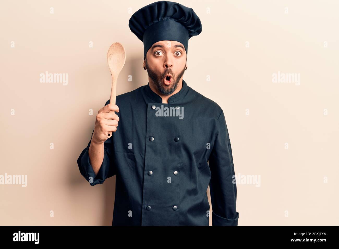 Young hispanic man wearing cooker uniform holding spoon scared and ...