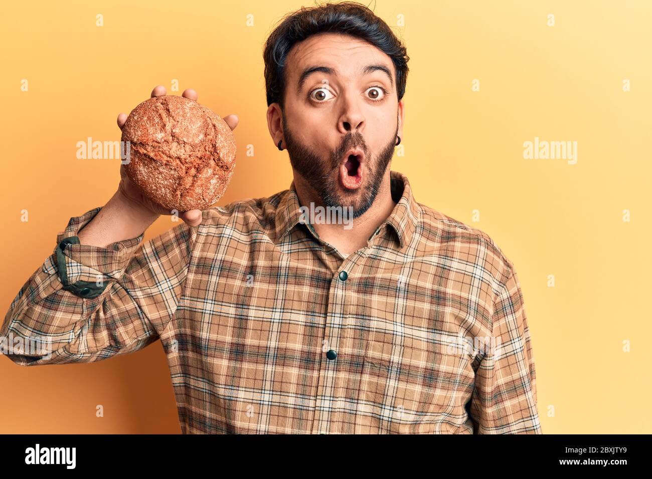 Young hispanic man holding bread scared and amazed with open mouth for ...