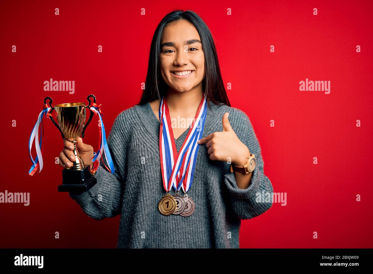 Young beautiful champion asian woman holding trophy wearing medals over ...