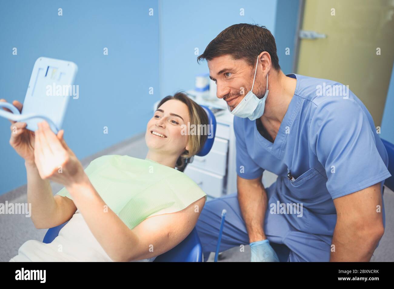 Dentist shows the patient the results of treatment with a mirror, examinating teeth with dental ...