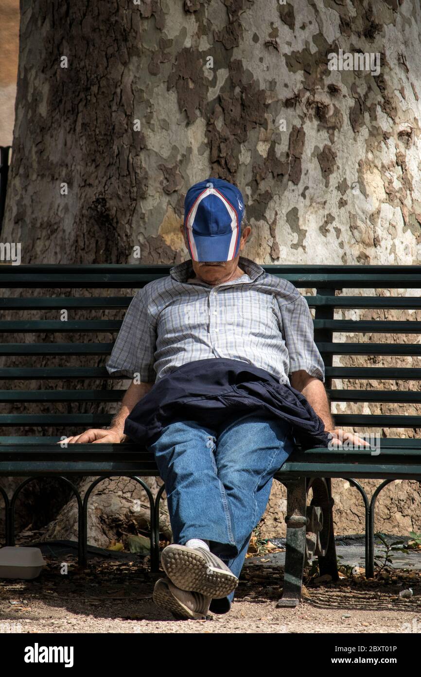 A man is dozing in the sun on a park bench with his cap down on his ...