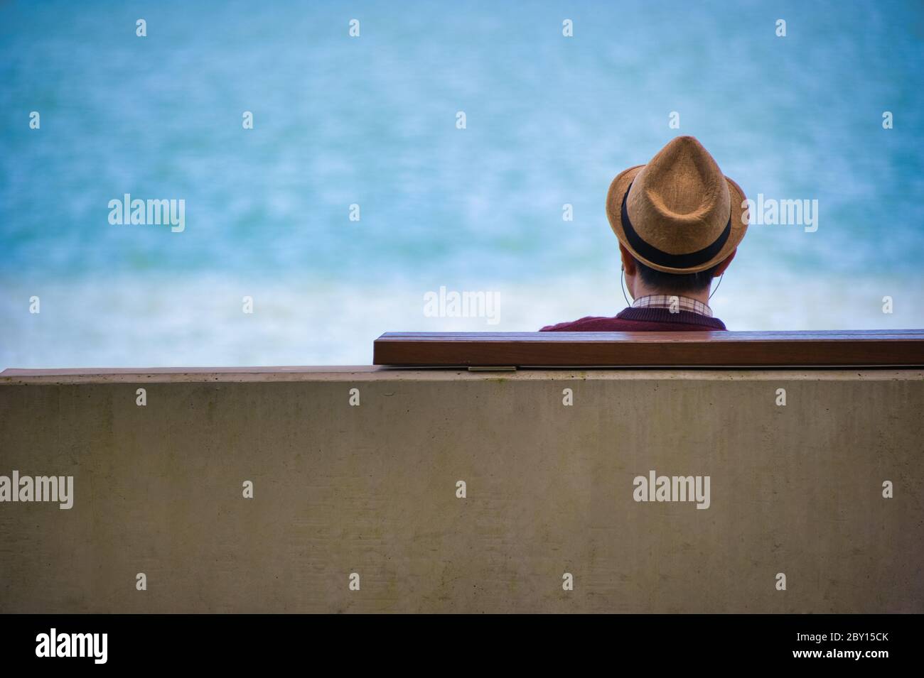 Tourist with a fedora sits relaxed on a seaside park bench looking out ...