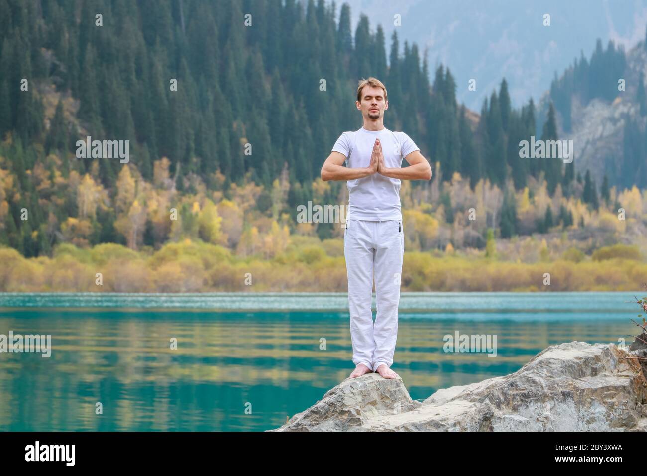 A young man in white practices yoga in the mountains. Pose Samasthiti ...