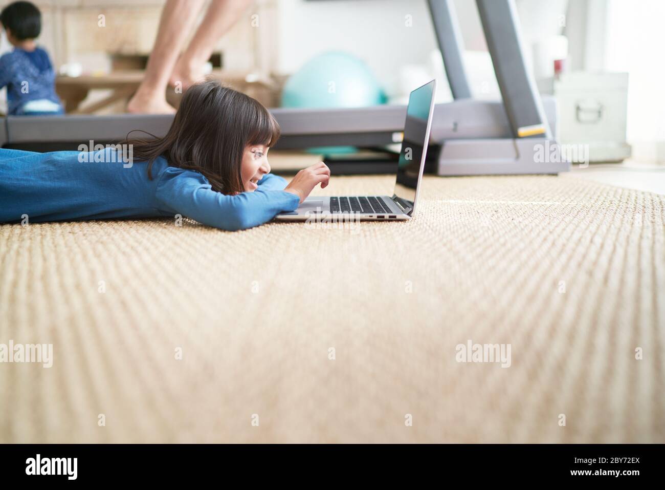 Girl using laptop on living room floor next to father on treadmill ...