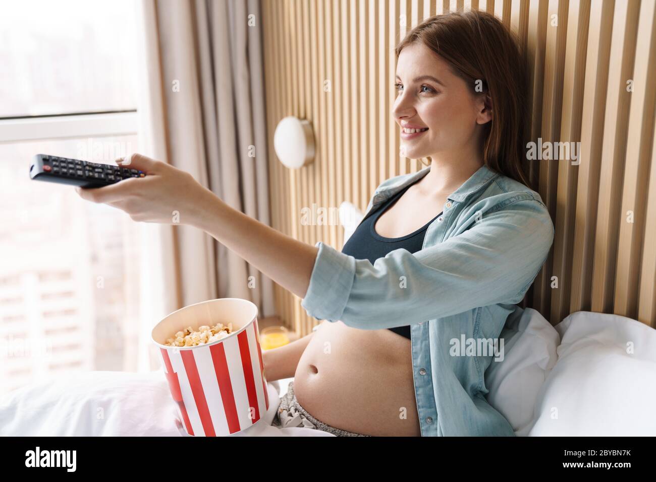 Photo of smiling pregnant woman eating popcorn and using remote control ...