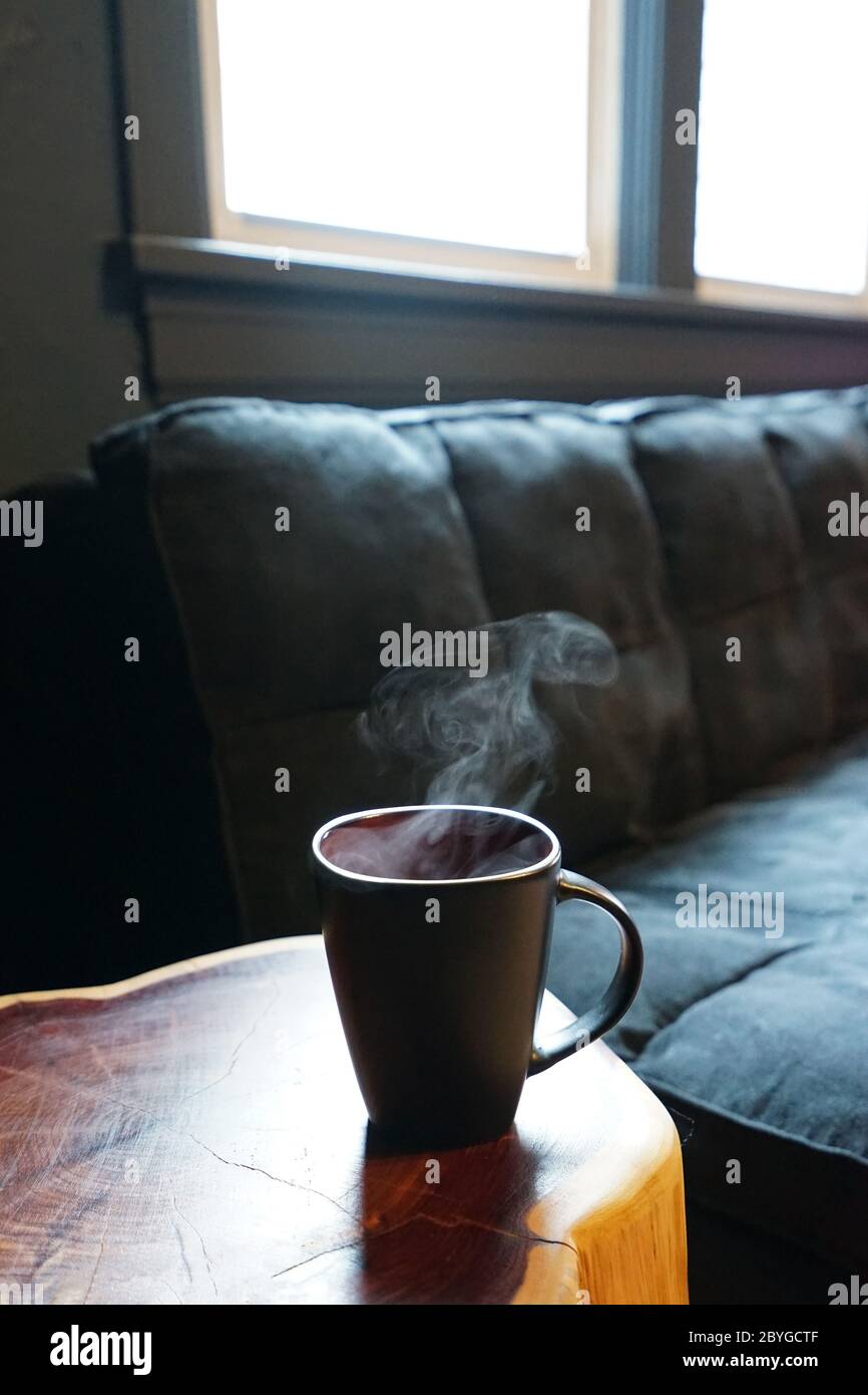 A mug of hot coffee on wooden stool table with cushion sofa on the back ...