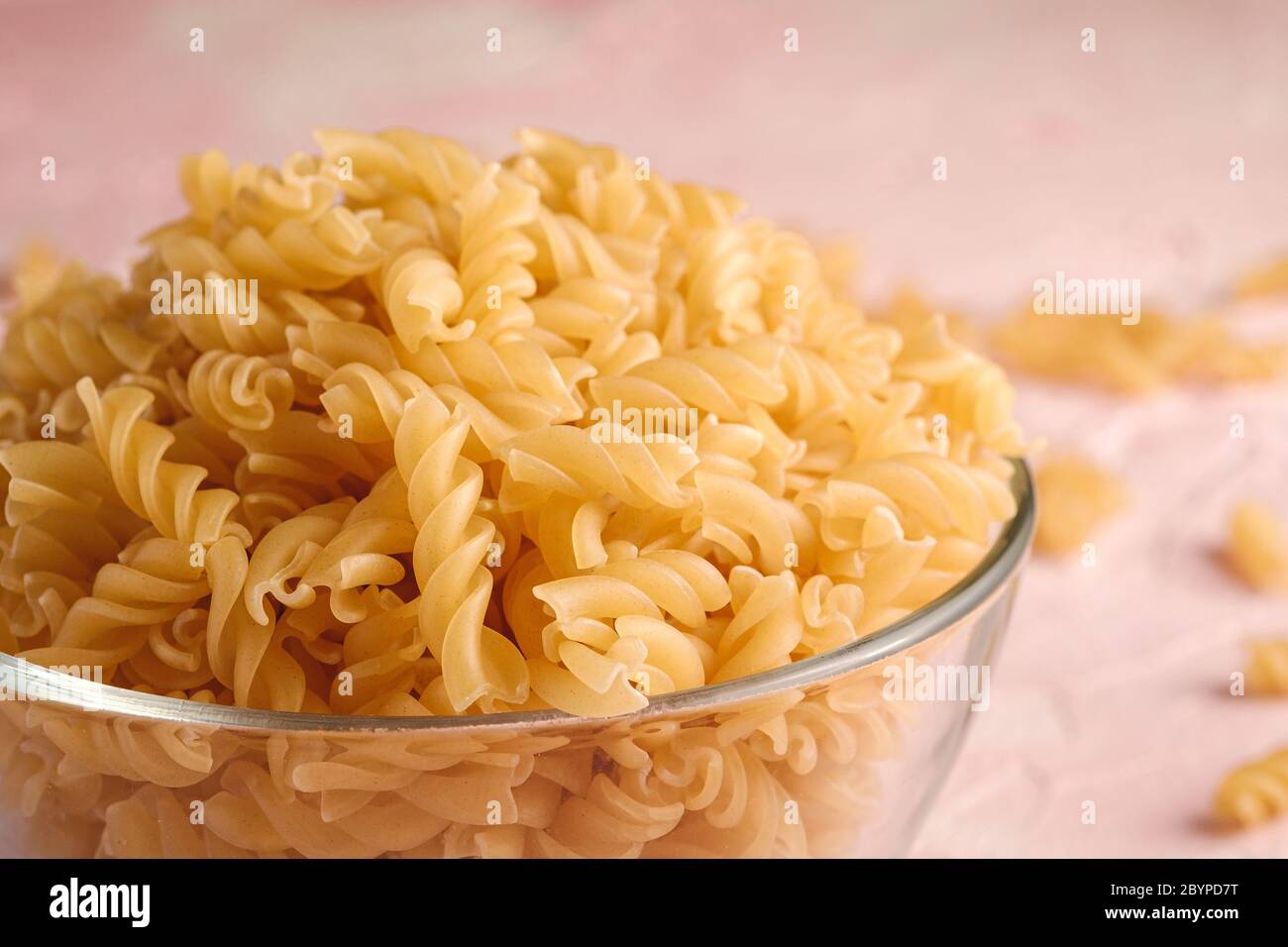 Raw wheat golden spiral pasta in glass bowl on textured pink background ...