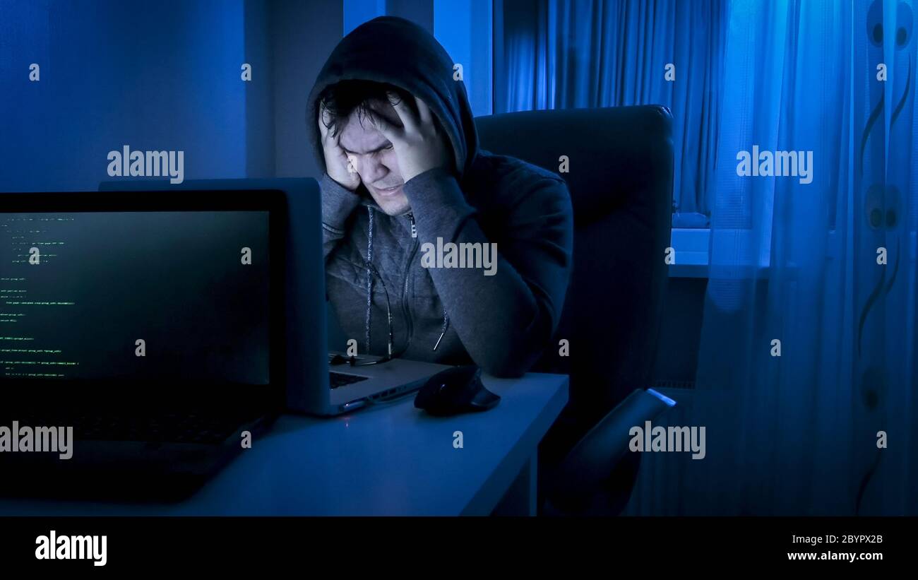 Angry male hacker in hood holding hands on his forehead while working ...