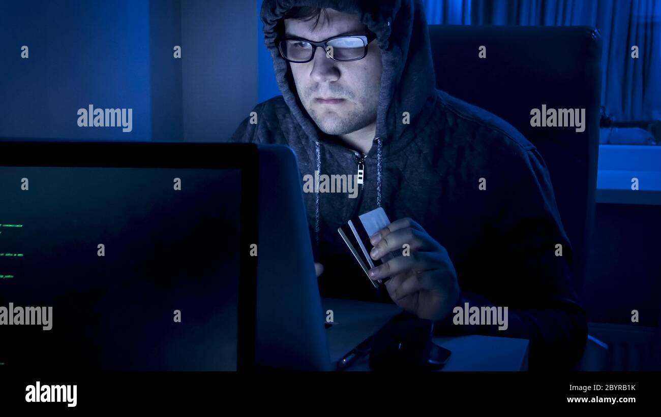 Portrait of hacker holding credit cards working on computer at night ...