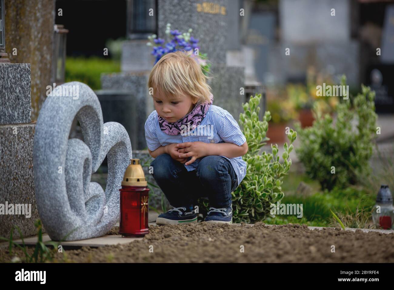 Little toddler boy, sitting on a grave in cemetery, sad and lonely ...