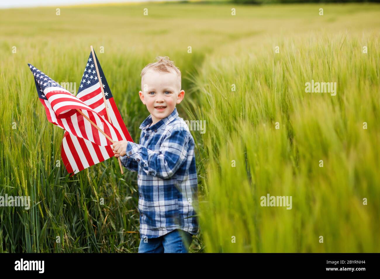A cute blond boy is standing in a field with an American flag in his ...