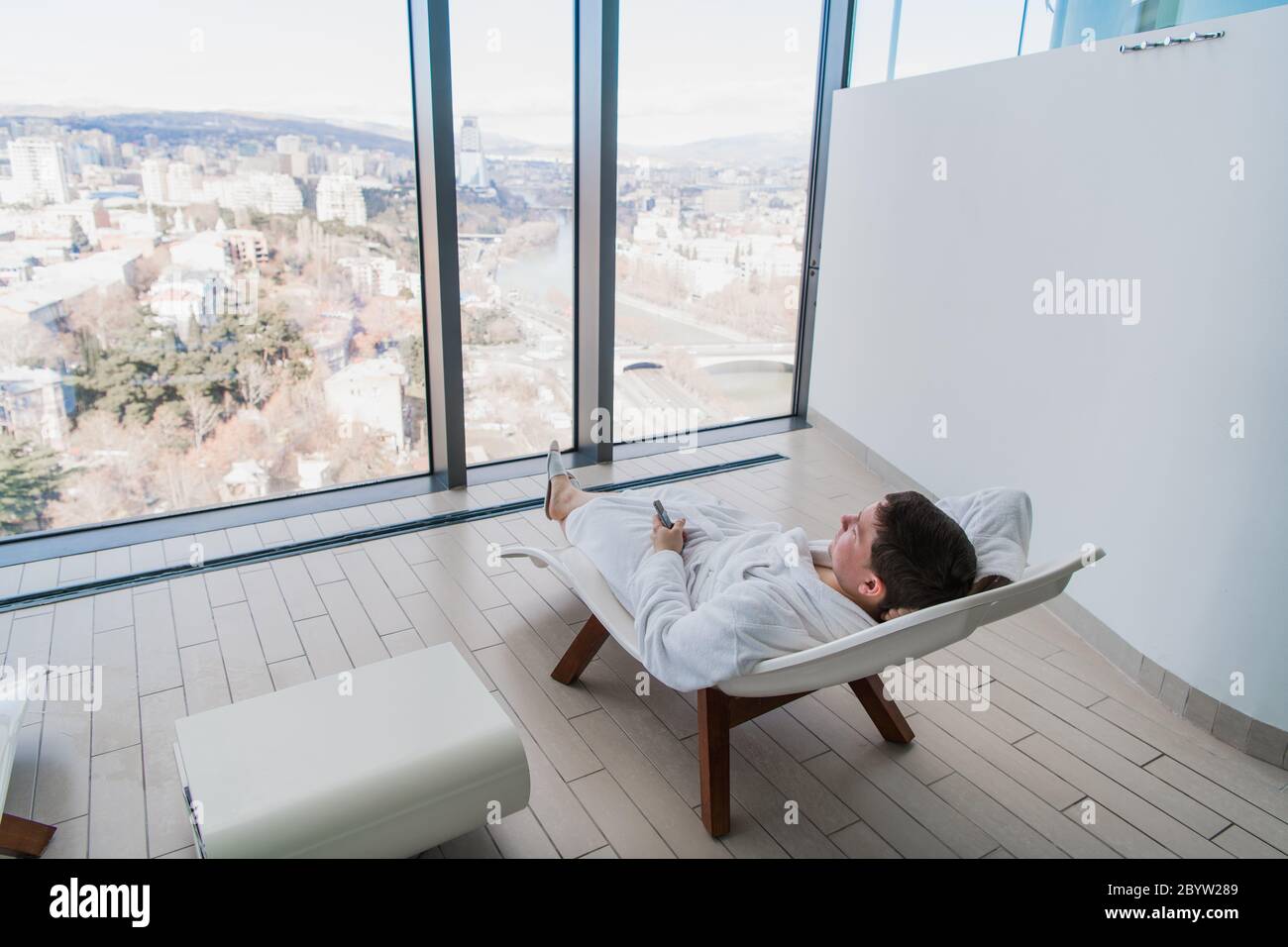 Young man resting on modern deck chair in front of big window with city ...