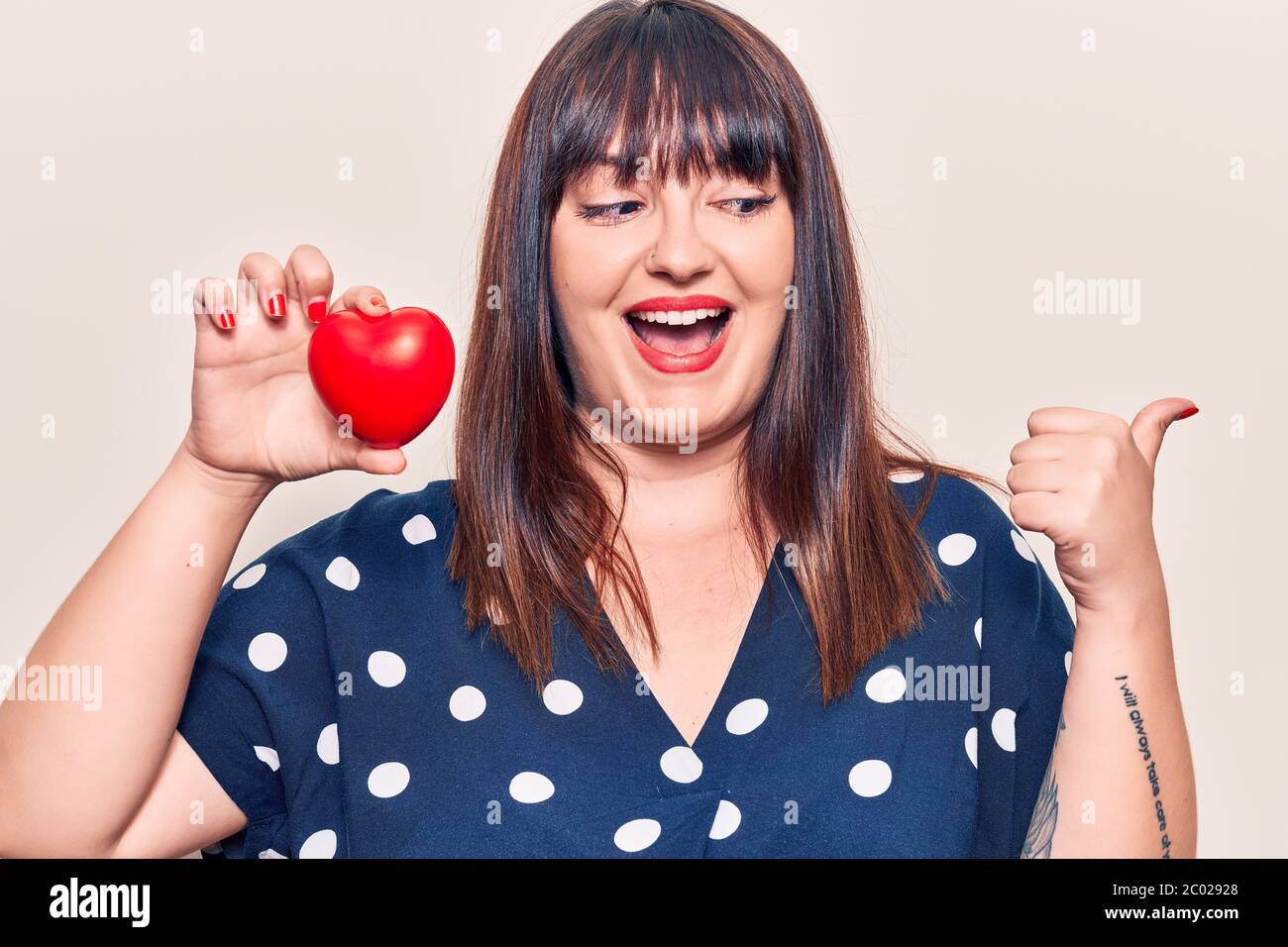 Young plus size woman holding heart pointing thumb up to the side ...
