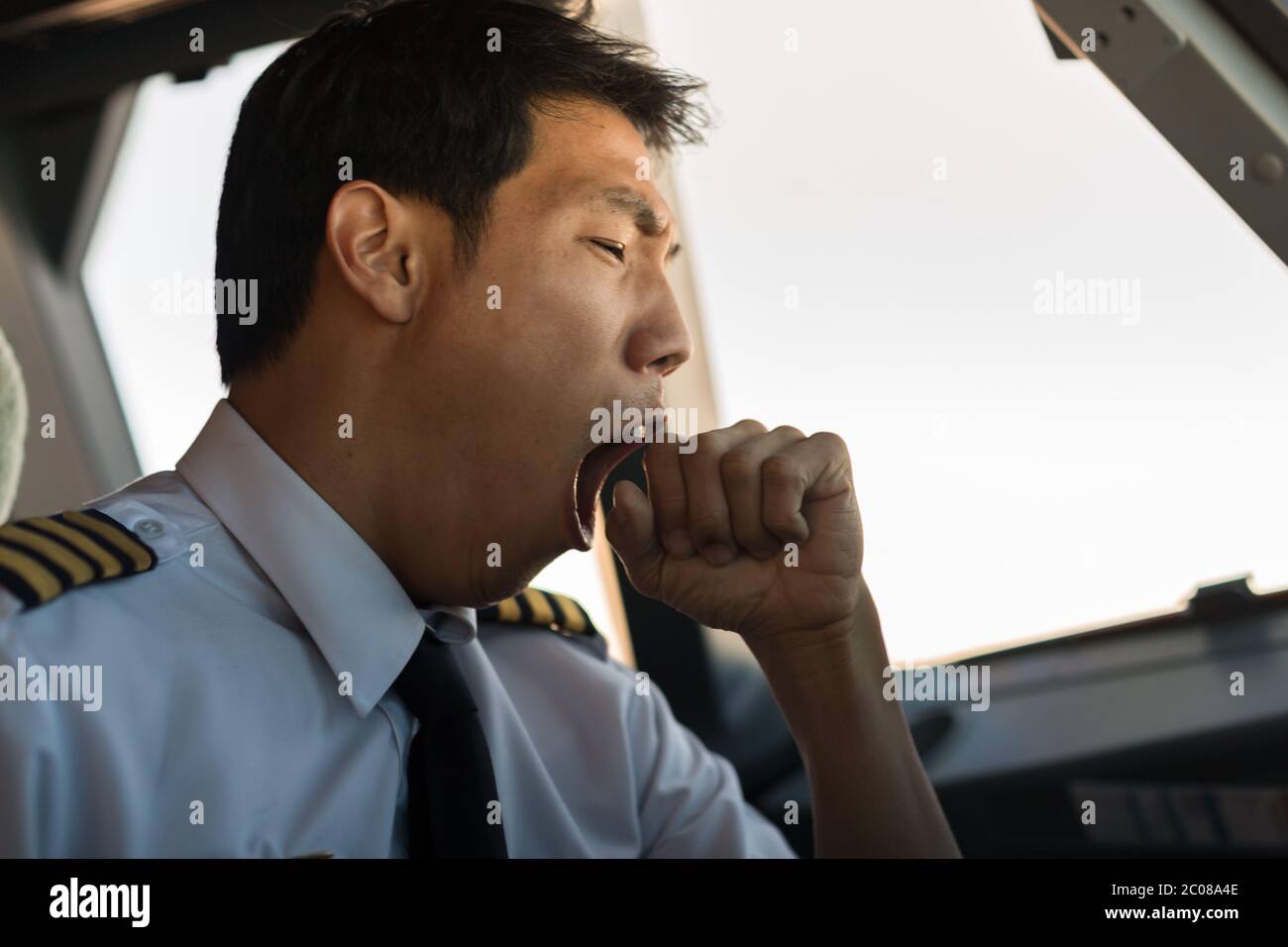 A male captain pilot sitting in the cockpit yawning while flying an ...