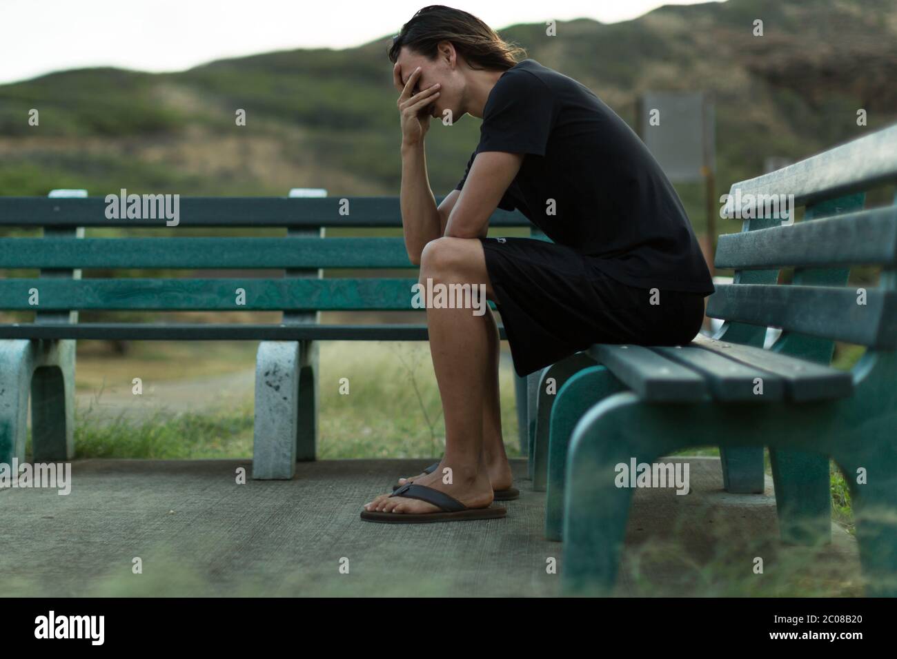A young man sitting on the park bench outside, stressed out covering ...
