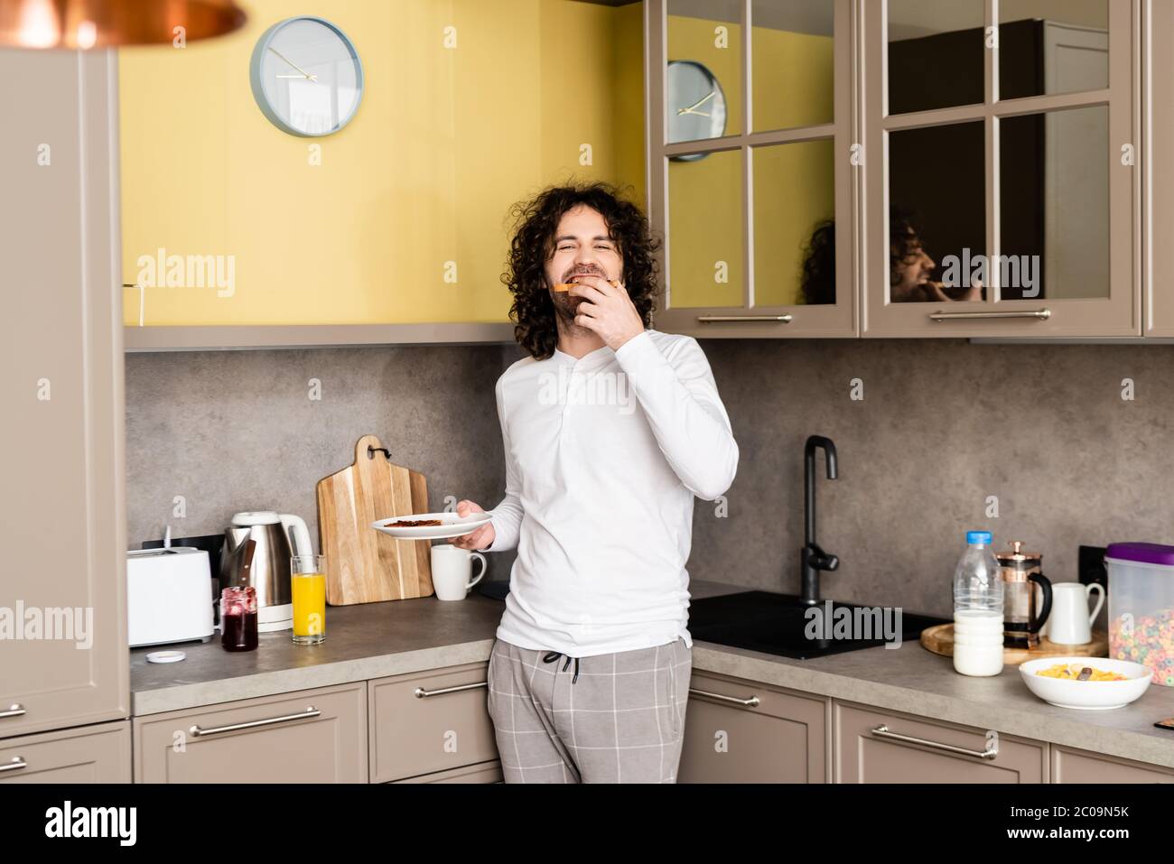 joyful, curly man eating delicious toast with jam while looking at ...